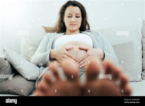 Description: Frontal view of woman lying on sofa gently holding her belly in last months of ...